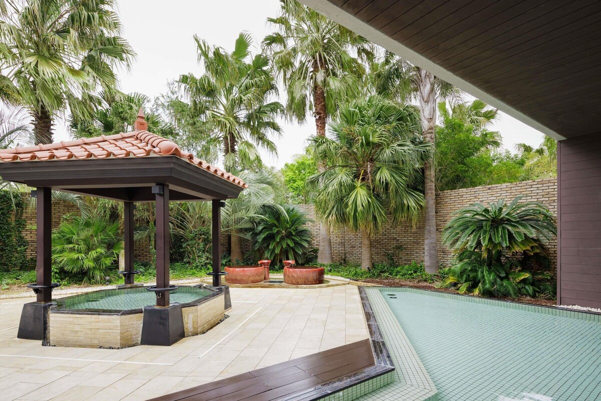 Área de piscina com um gazebo, palmeiras e um muro de tijolos.