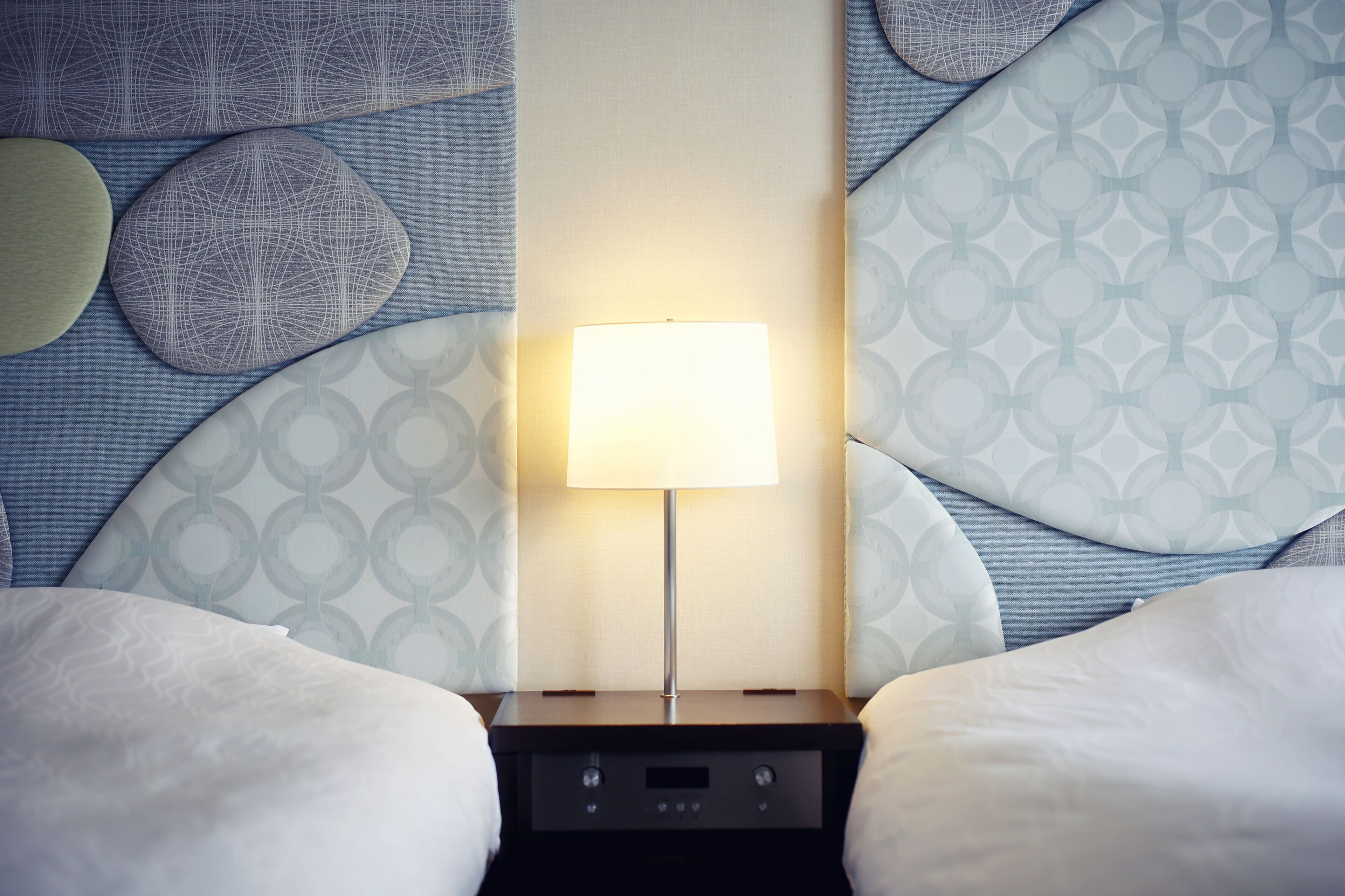 Cama bem arrumada com uma luminária moderna ao lado, um painel de controle e uma parede estampada.