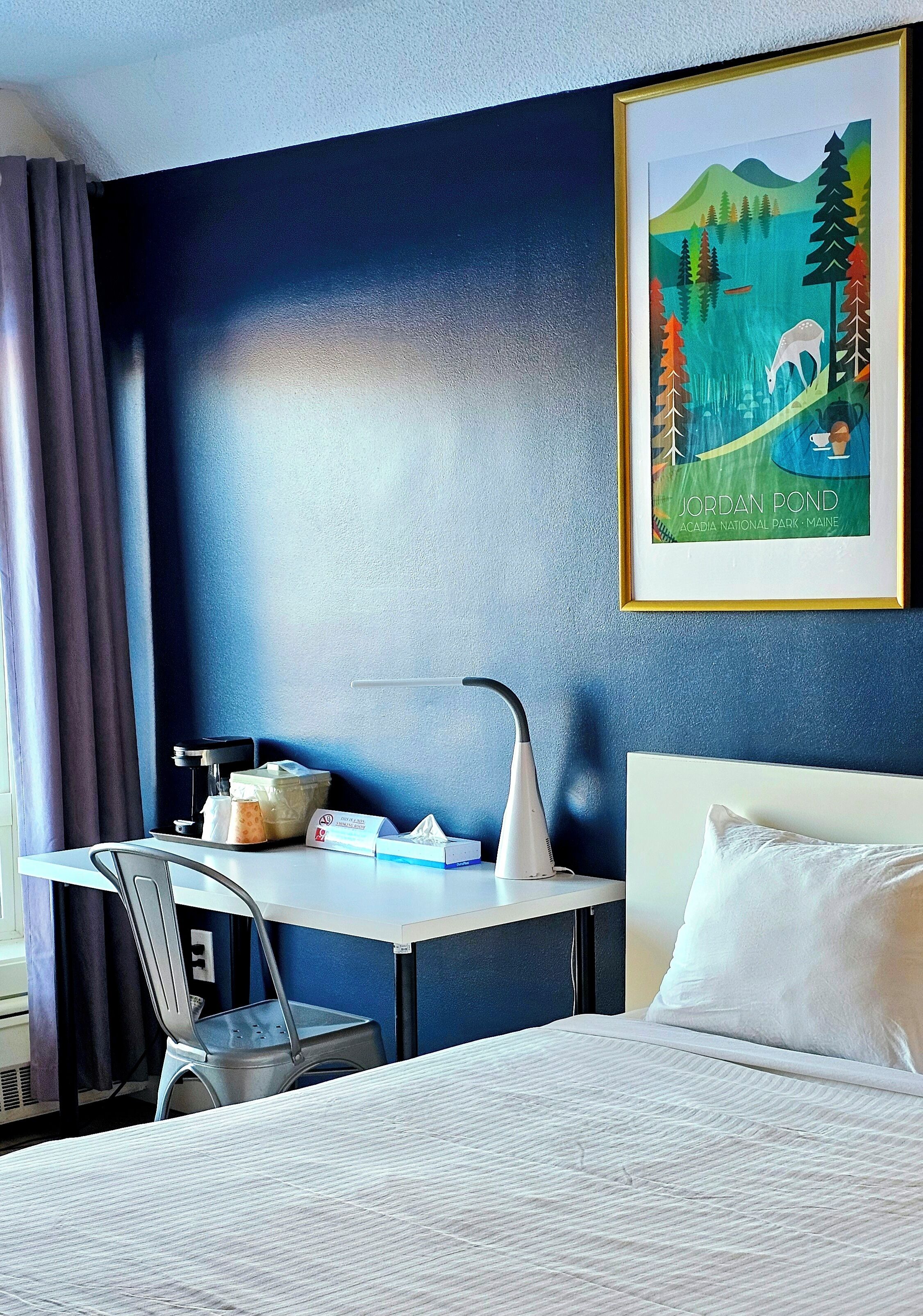 Quarto de hotel com cama, escrivaninha com cadeira, cafeteira, moldura de quadro e janela com cortinas.