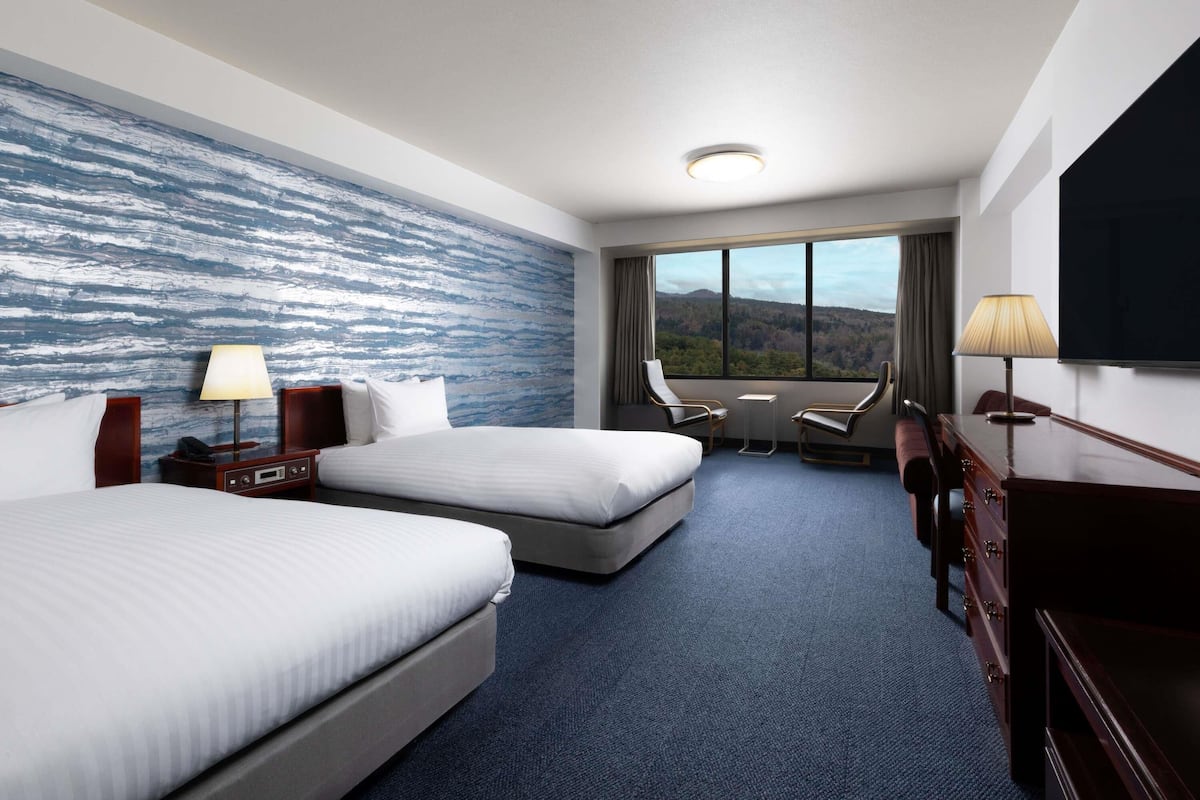 Quarto de hotel com duas camas, uma escrivaninha, uma cadeira, uma televisão e uma janela grande com vista para as montanhas.