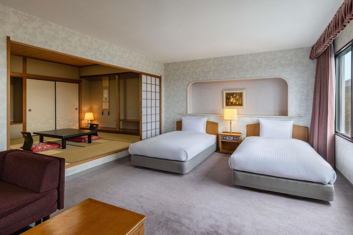 Quarto de hotel com duas camas, um sofá, uma mesinha e um quadro na parede, além de uma porta deslizante tradicional.