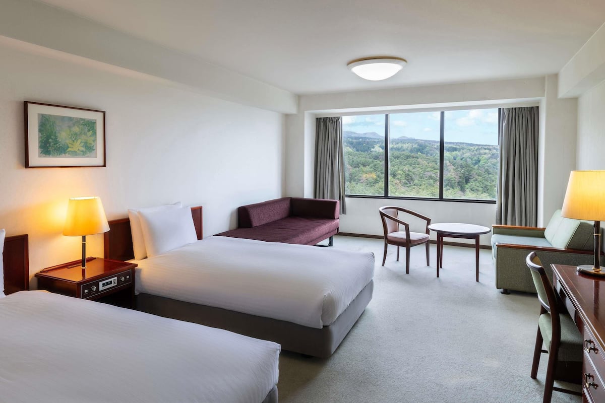 Quarto de hotel com duas camas, uma escrivaninha, uma cadeira e vista para o exterior.