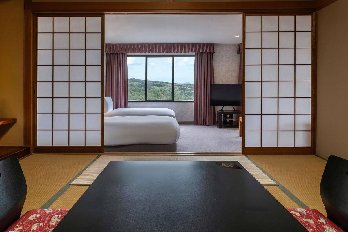 Quarto em estilo japonês tradicional, com tatames, cama, televisão e vista para uma paisagem arborizada.