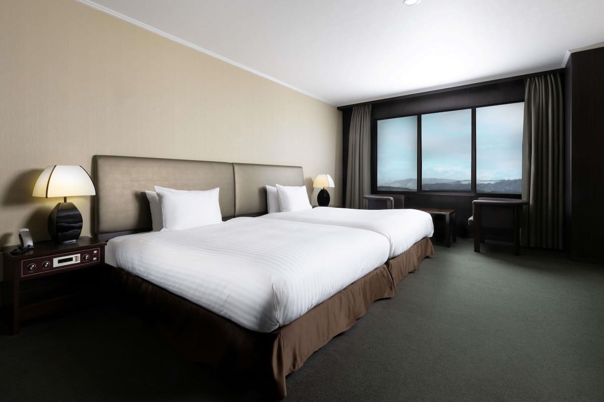 Quarto de hotel com duas camas, uma janela grande, uma escrivaninha e uma cadeira.