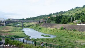 Um rio serpenteando sob uma ponte, com vegetação nas margens e uma cerca ao primeiro plano.