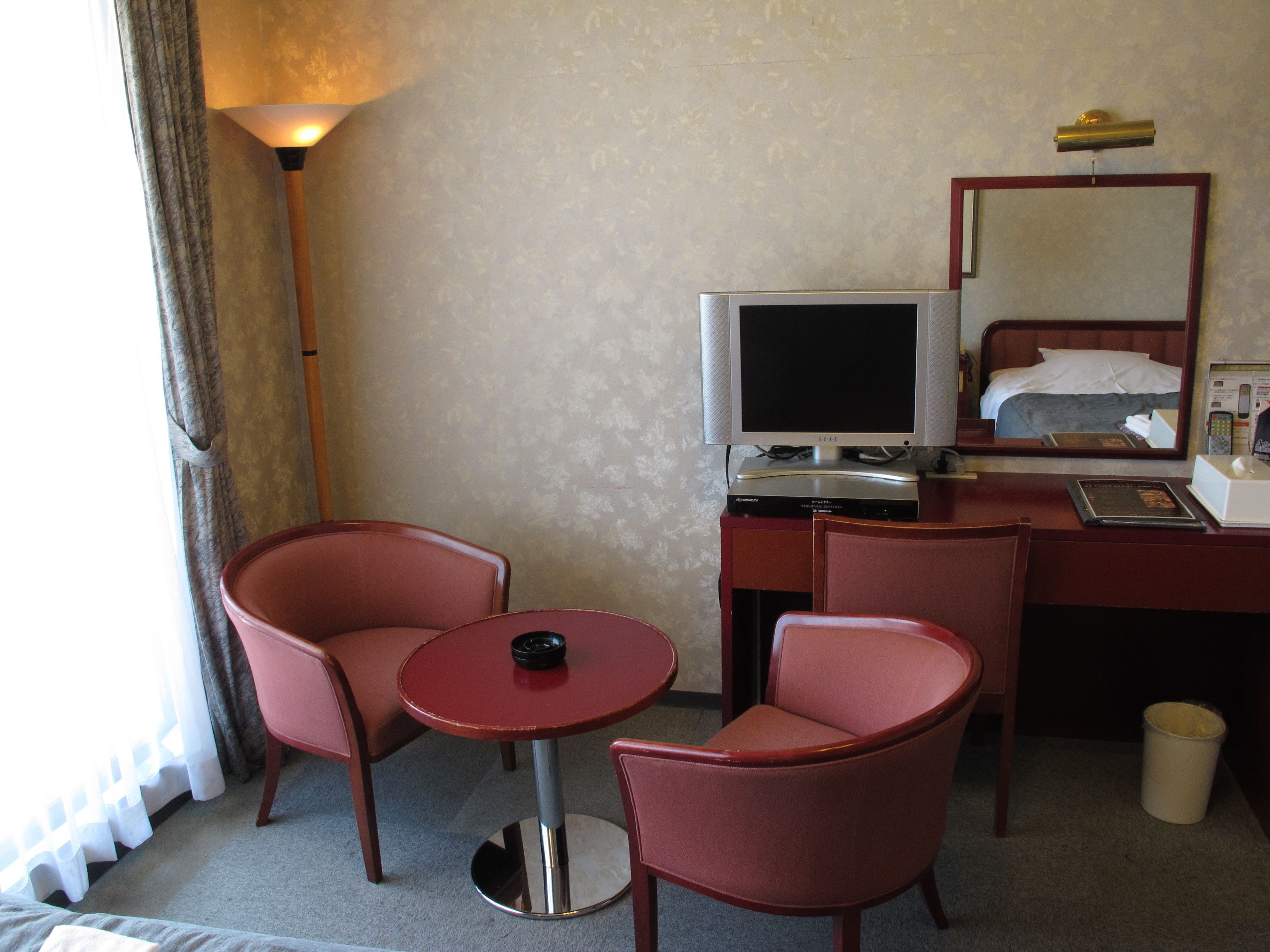 Quarto de hotel com escrivaninha, duas cadeiras, televisão, abajur, cama e lixeira.