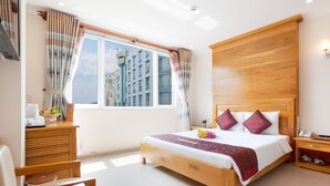 Quarto de hotel com uma cama grande, cabeceira de madeira, uma escrivaninha com telefone e uma janela com cortinas.