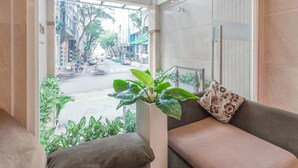 Um saguão moderno de hotel com vista para uma rua movimentada, uma grande planta em vaso e uma área de estar com sofá e almofada estampada.