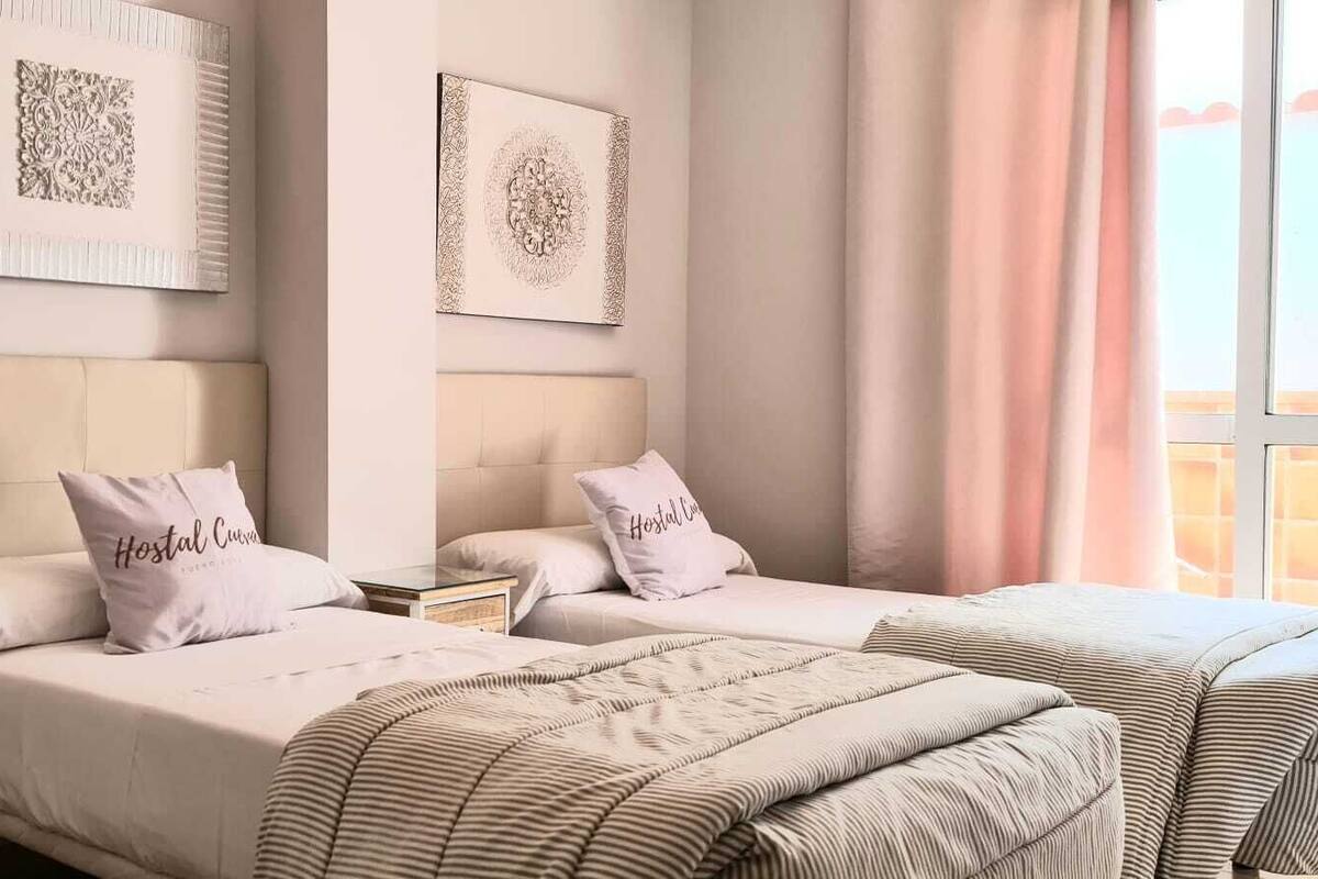 Quarto de hotel com duas camas, cada uma com um travesseiro rotulado "Hostal Casa". Há obras de arte fixadas na parede acima das camas, uma mesa de cabeceira com abajur e uma janela com cortinas.