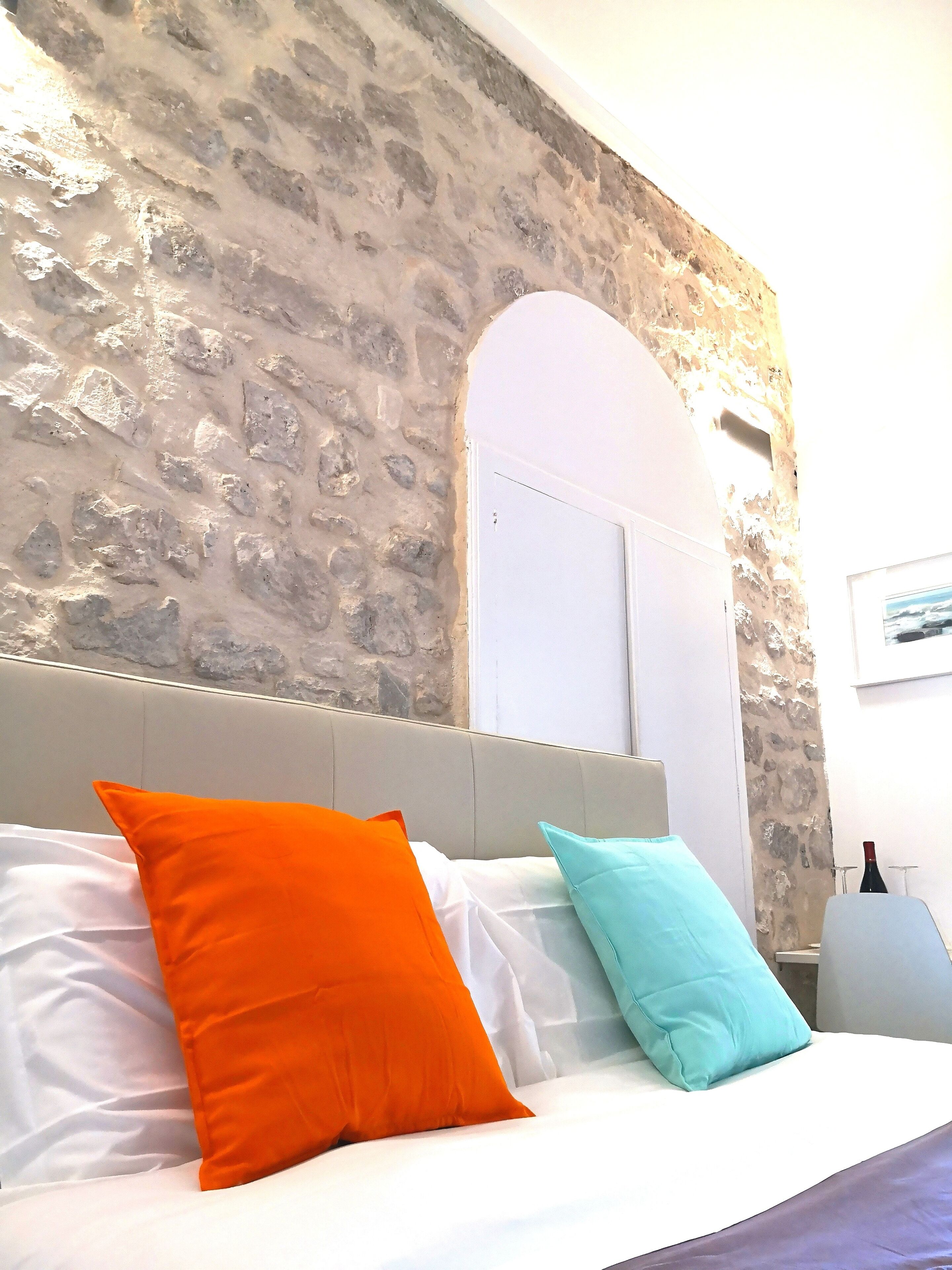 Uma cama com almofadas brancas e laranja, uma parede de pedra e uma porta branca.