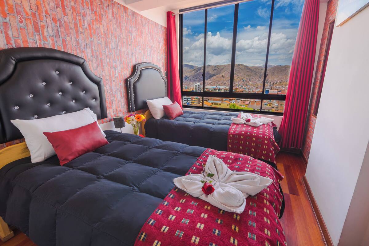 Quarto de hotel com duas camas, uma janela grande com vista para as montanhas e parede com padrão de tijolos.