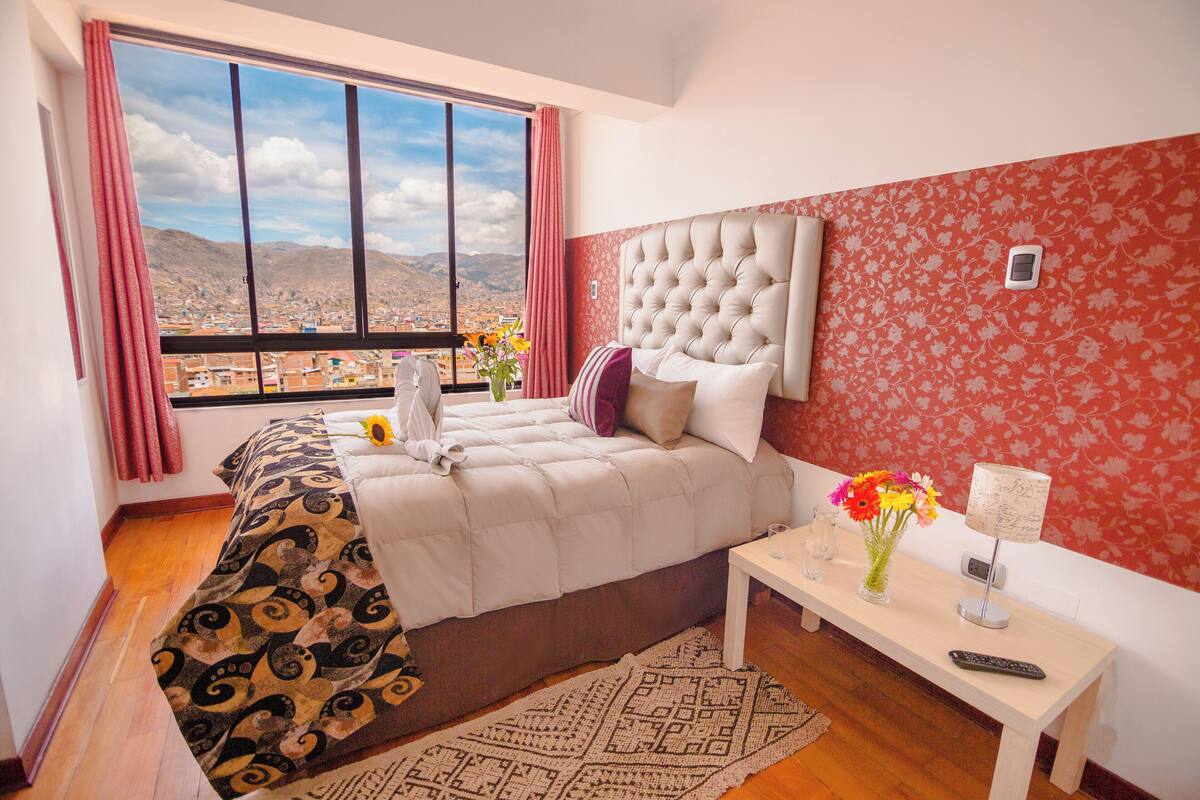 Quarto com uma grande janela, cama com roupa de cama estampada, mesa de cabeceira com abajur e um vaso, e vista para uma paisagem montanhosa.