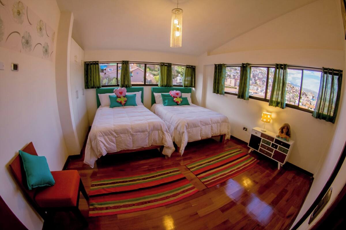 Quarto com duas camas, uma mesa de cabeceira, uma cadeira e vista para a cidade através das janelas.