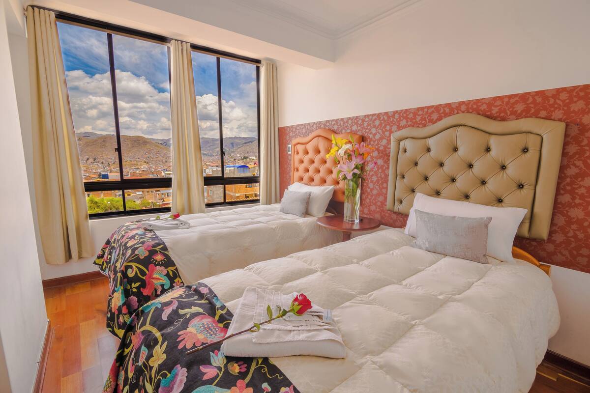 Quarto de hotel com duas camas, uma janela grande com vista para a cidade e um edredom estampado com motivos florais.