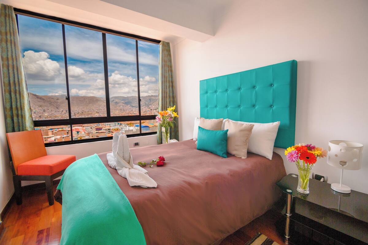 Quarto de hotel com uma grande janela, cama com cabeceira verde-água, uma cadeira, mesa de cabeceira com abajur e um vaso com flores.