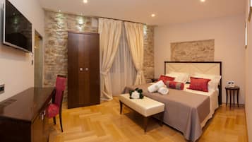 Quarto de hotel com cama, um banquinho, televisão, guarda-roupa e parede de pedra.