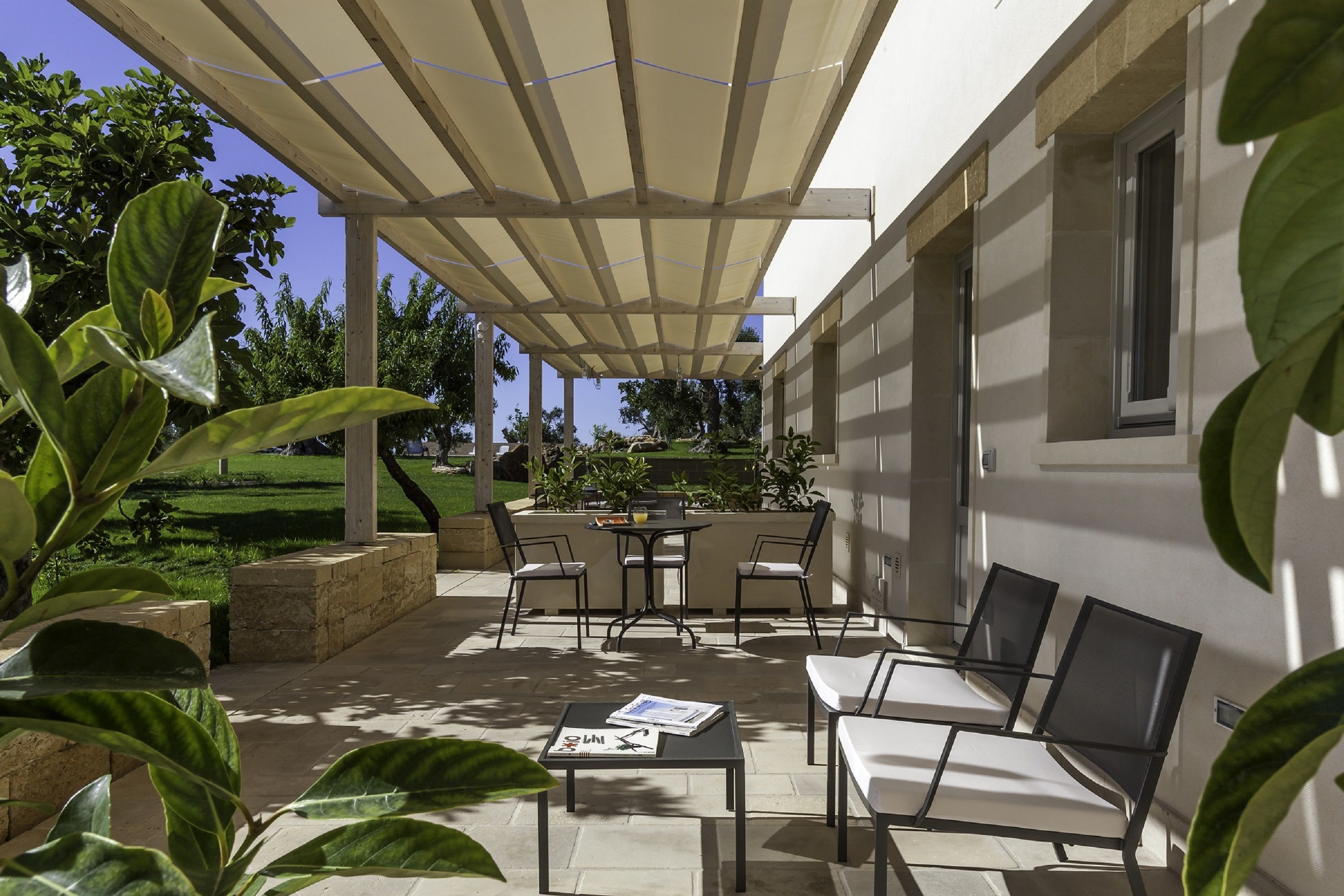Um pátio externo com mesa e cadeiras, cercado por plantas em vasos e uma pérgola.