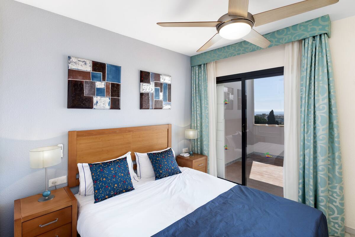 Quarto de hotel com cama, mesas de cabeceira, ventoinha no teto, obras de arte nas paredes e vista através da porta de vidro.