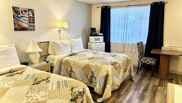 Quarto de hotel com duas camas, uma escrivaninha, uma cadeira, janela com cortinas, luminária e uma obra de arte na parede.
