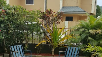 Uma área externa gramada com duas cadeiras de plástico azuis e uma branca, uma pequena árvore e um edifício com telhado cinzento ao fundo.