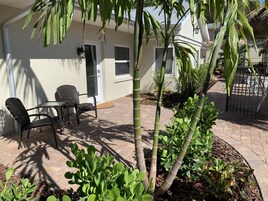 Uma área de pátio com mobiliário externo, uma mesa pequena e cadeiras. Há plantas e um portão de metal ao fundo.