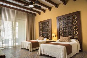 Quarto de hotel com duas camas, uma janela grande com cortinas, um ventilador de teto e um painel decorativo de madeira na parede.