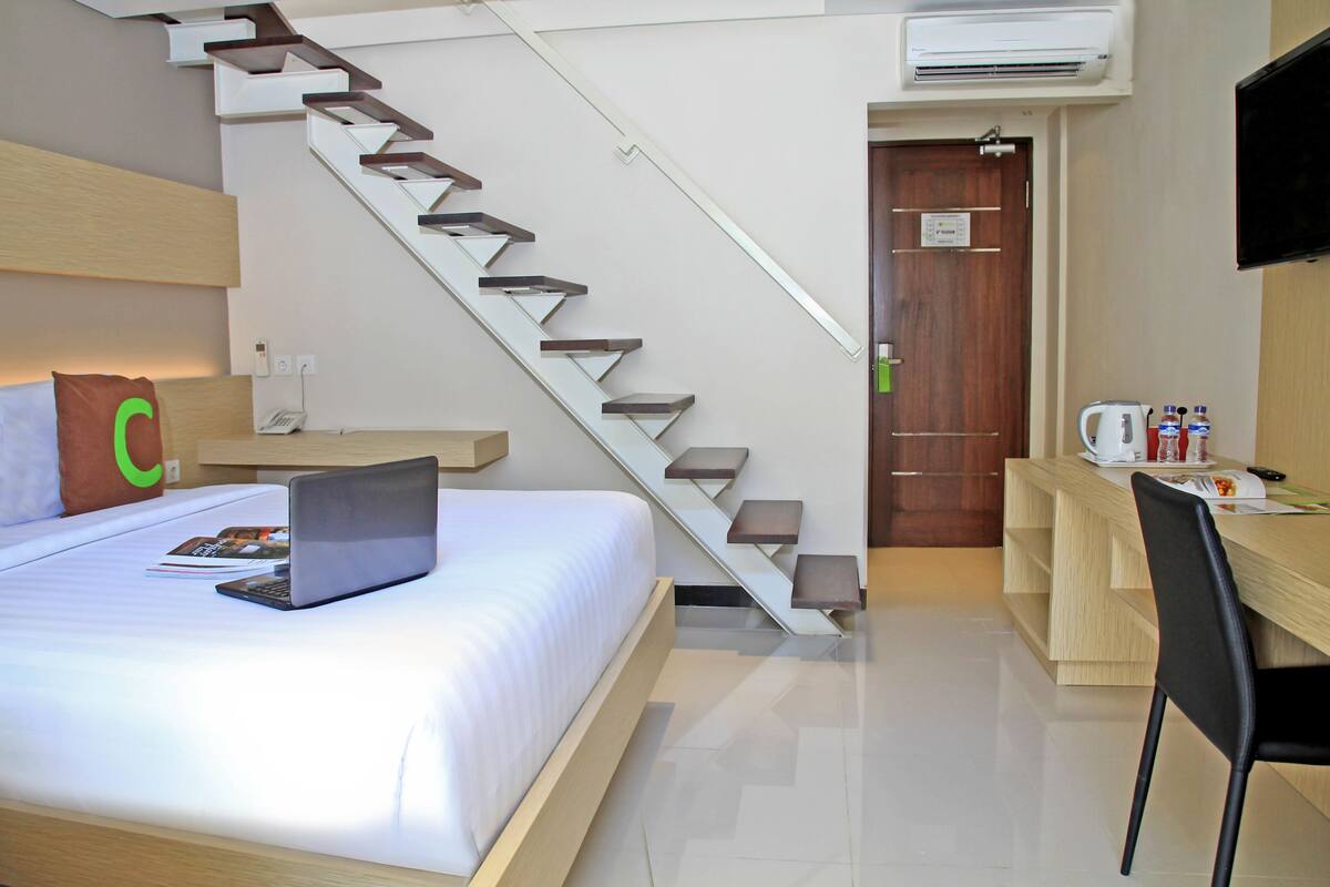 Quarto de hotel moderno com uma cama, um laptop, uma escrivaninha com uma cadeira, uma escada de madeira e uma televisão.
