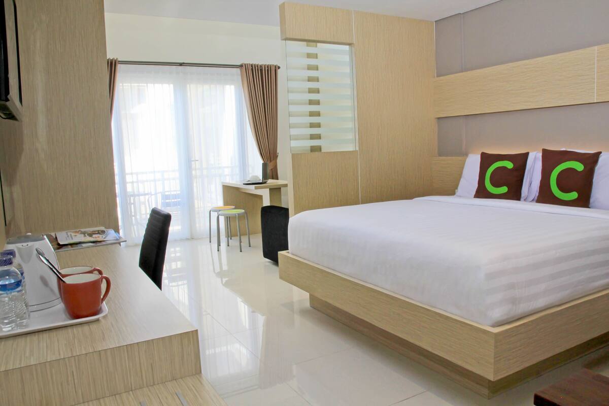 Quarto de hotel moderno com uma cama grande, uma escrivaninha com cadeira, uma mesinha e uma janela com cortinas.