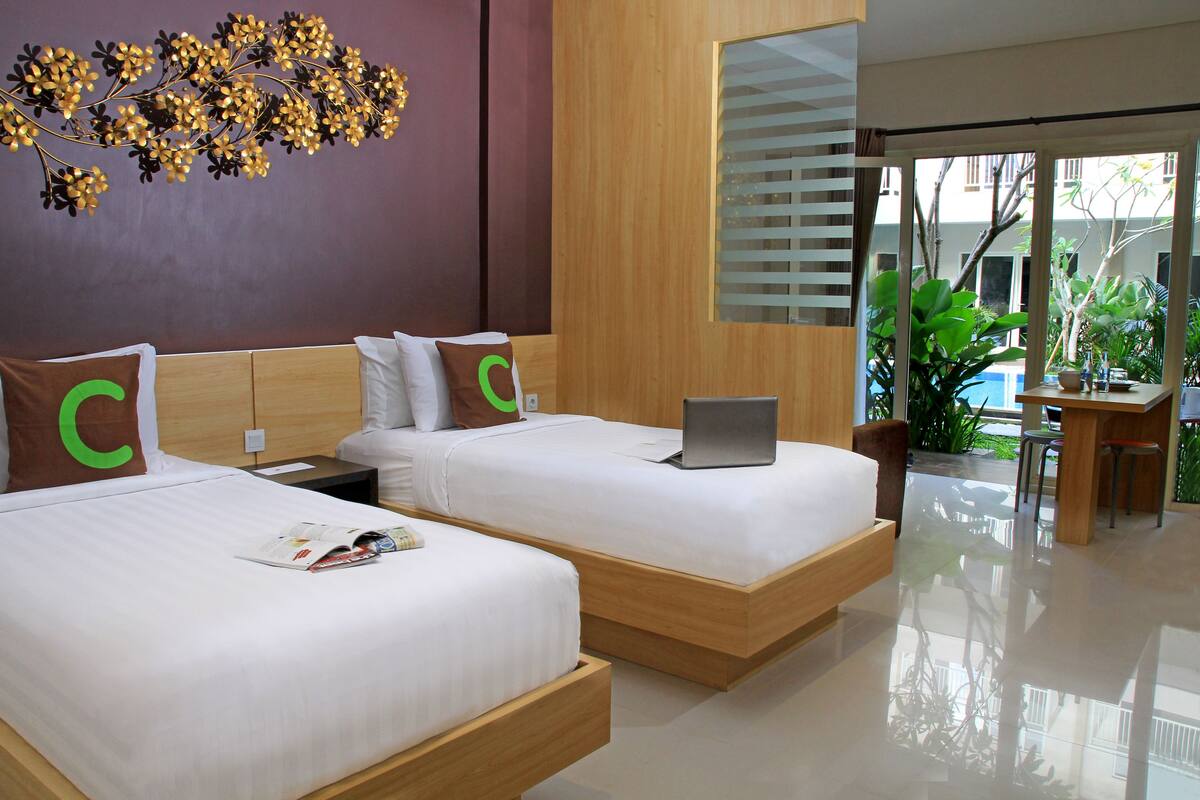 Quarto de hotel moderno com duas camas, um laptop em uma delas e vista para a área externa através de grandes janelas.