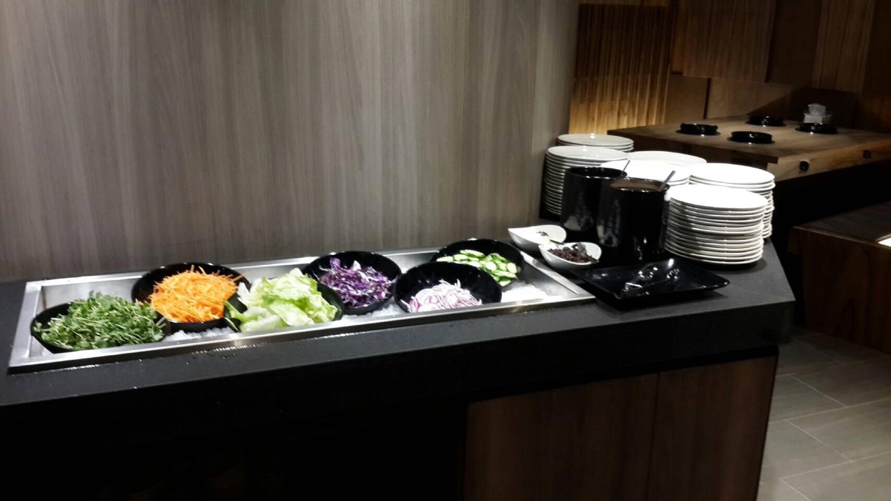 Buffet com diversos alimentos em recipientes, pratos empilhados no balcão e um armário de madeira ao fundo.