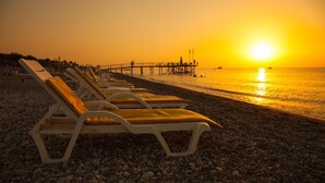 Uma praia com fileiras de espreguiçadeiras viradas para o mar, um píer que se estende na água e um pôr do sol ao fundo.