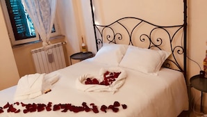 Cama bem arrumada com lençóis brancos, um coração decorativo feito de pétalas de rosa e um frasco de perfume na mesa de cabeceira.