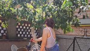 Uma pessoa ao lado de uma bicicleta turquesa com cesto, em frente a um edifício com parede estampada e uma árvore.