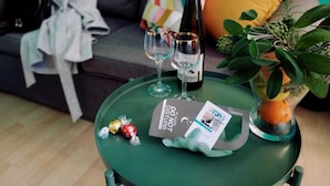 Uma mesa de centro verde com uma garrafa de vinho, duas taças de vinho, uma revista e uma pequena planta.