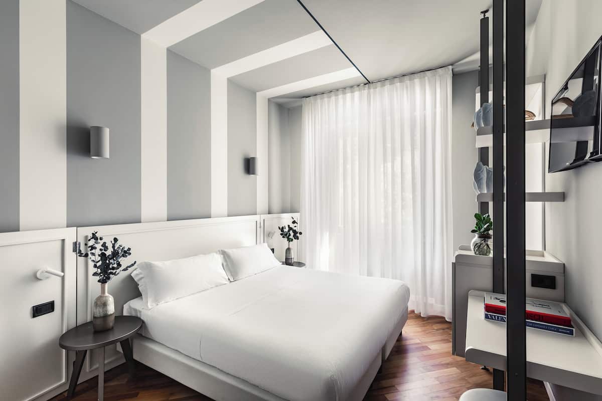 Quarto moderno com cama grande, mesas de cabeceira, mesinha lateral com um vaso e prateleira embutida para TV.