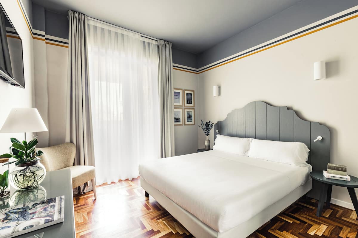 Quarto de hotel bem arrumado com uma cama grande, uma cadeira, uma mesa pequena com abajur e um vaso com flores.
