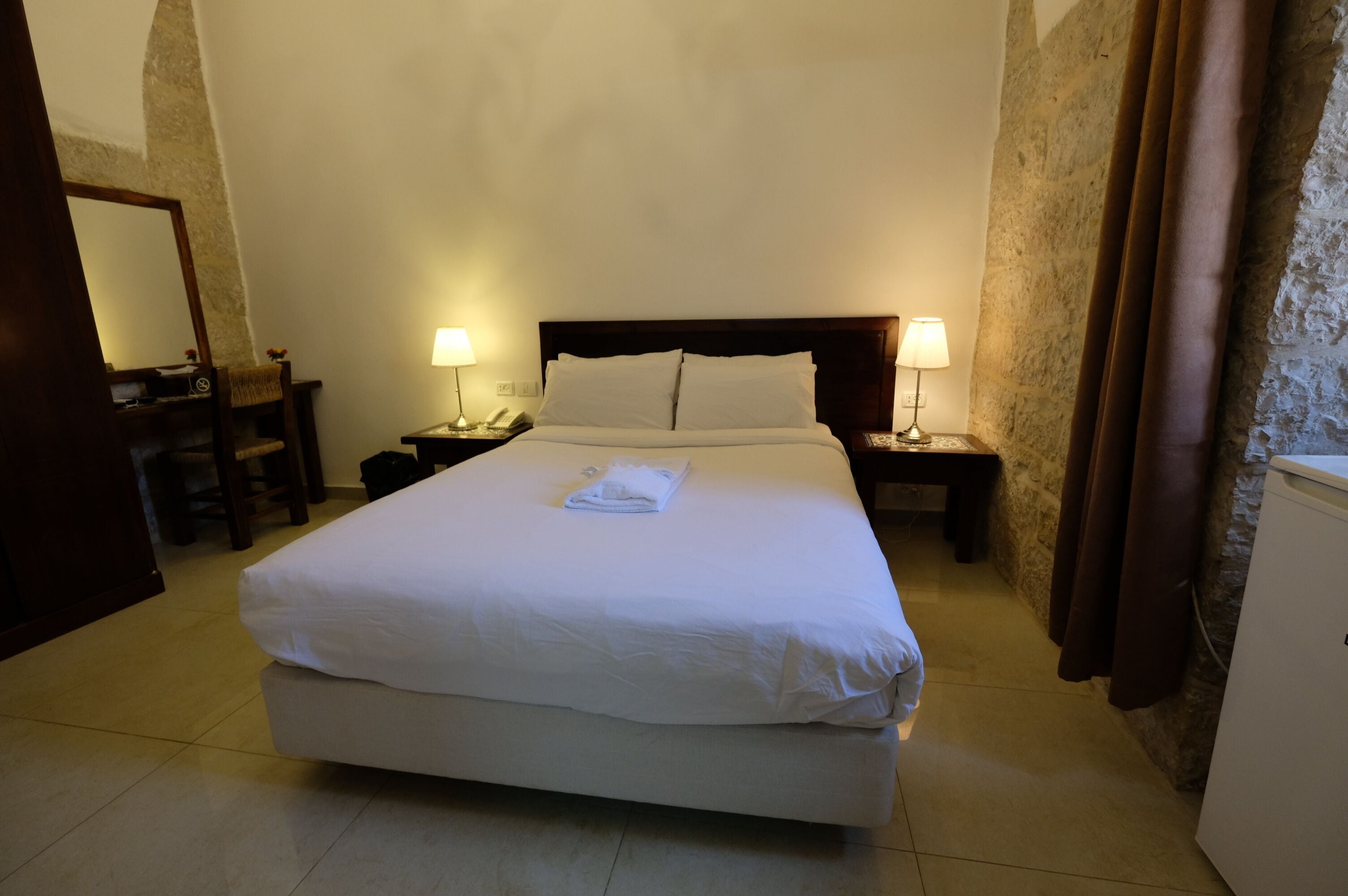 Cama bem arrumada com lençóis brancos e uma toalha dobrada, duas luminárias de cabeceira, mesa de cabeceira com telefone e uma pequena mesa com abajur, além de uma parede de pedra.