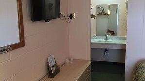 Quarto de hotel com televisão na parede, uma pequena mesa com telefone, espelho sobre a pia e uma cama com cobertura estampada.