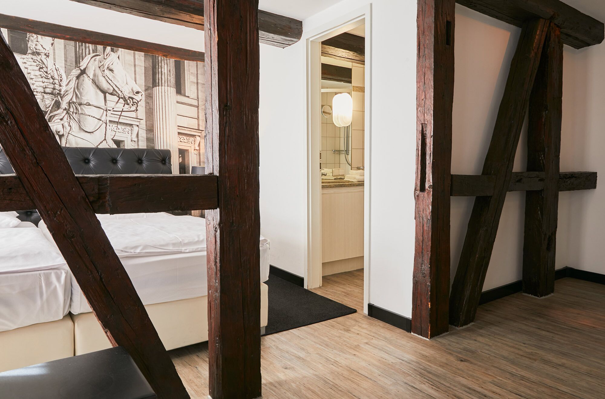 Quarto com cama, vigas de madeira e banheiro visível através de uma porta aberta.