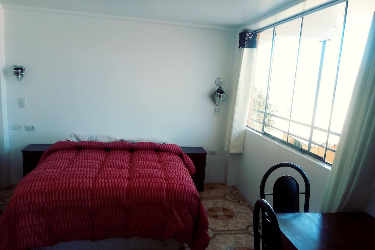 Quarto com cama, cadeira, mesa de cabeceira e janela com cortinas.