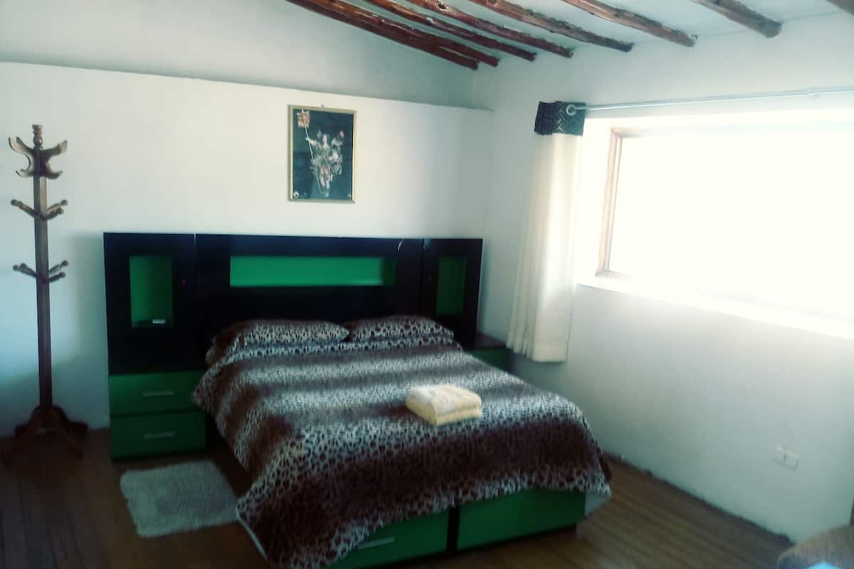 Quarto com cama verde, um cabideiro de madeira, quadro na parede e janela com cortinas.