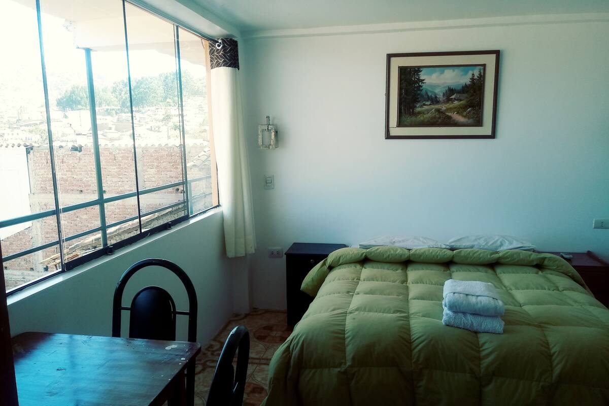 Quarto com cama, escrivaninha e cadeira. Janela com vista para edifícios e árvores.