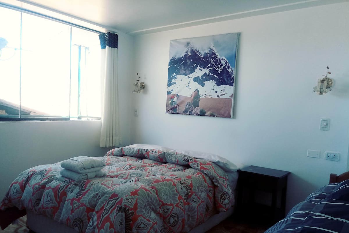Quarto com cama, mesa de cabeceira, janela com cortinas e um quadro na parede.