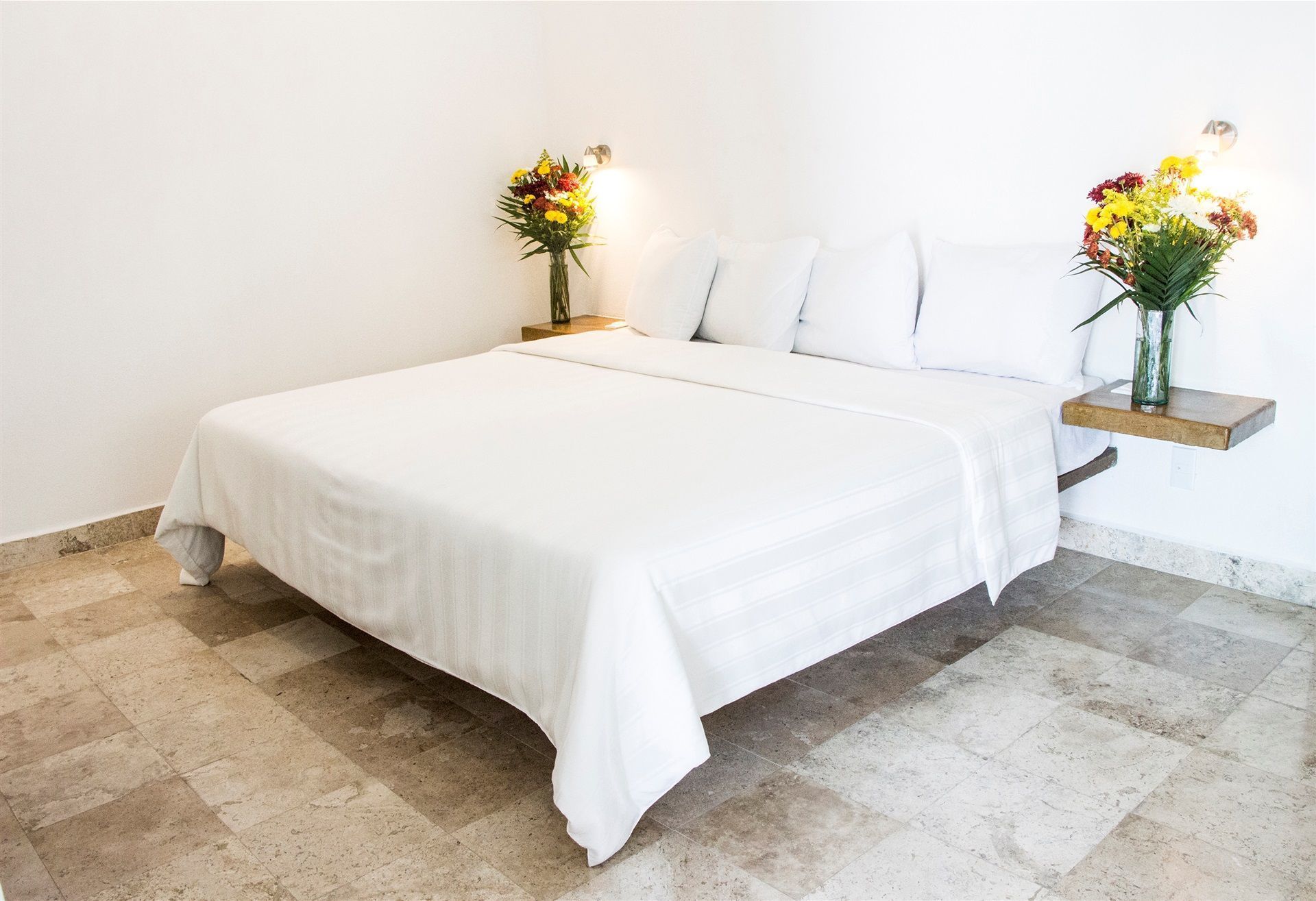 Uma cama bem arrumada com lençóis brancos, duas mesas de cabeceira com luminárias e uma pequena estante com um vaso de flores.