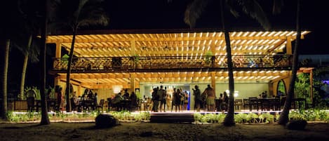 Um restaurante à beira-mar com um deque ao ar livre, palmeiras e chão de areia.