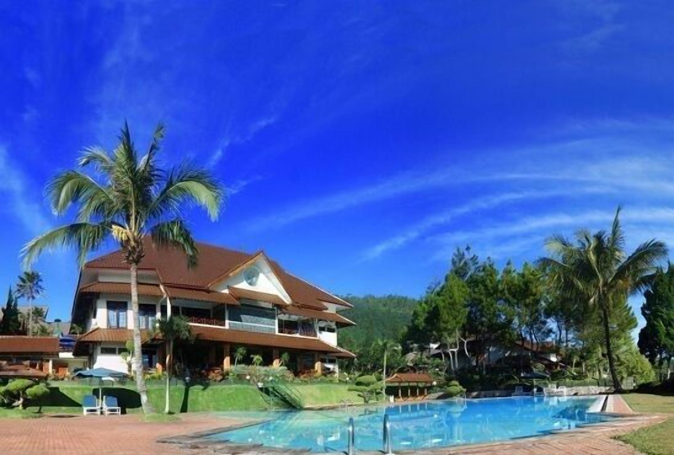 Hotel de dois andares com piscina, rodeado por palmeiras e vegetação.
