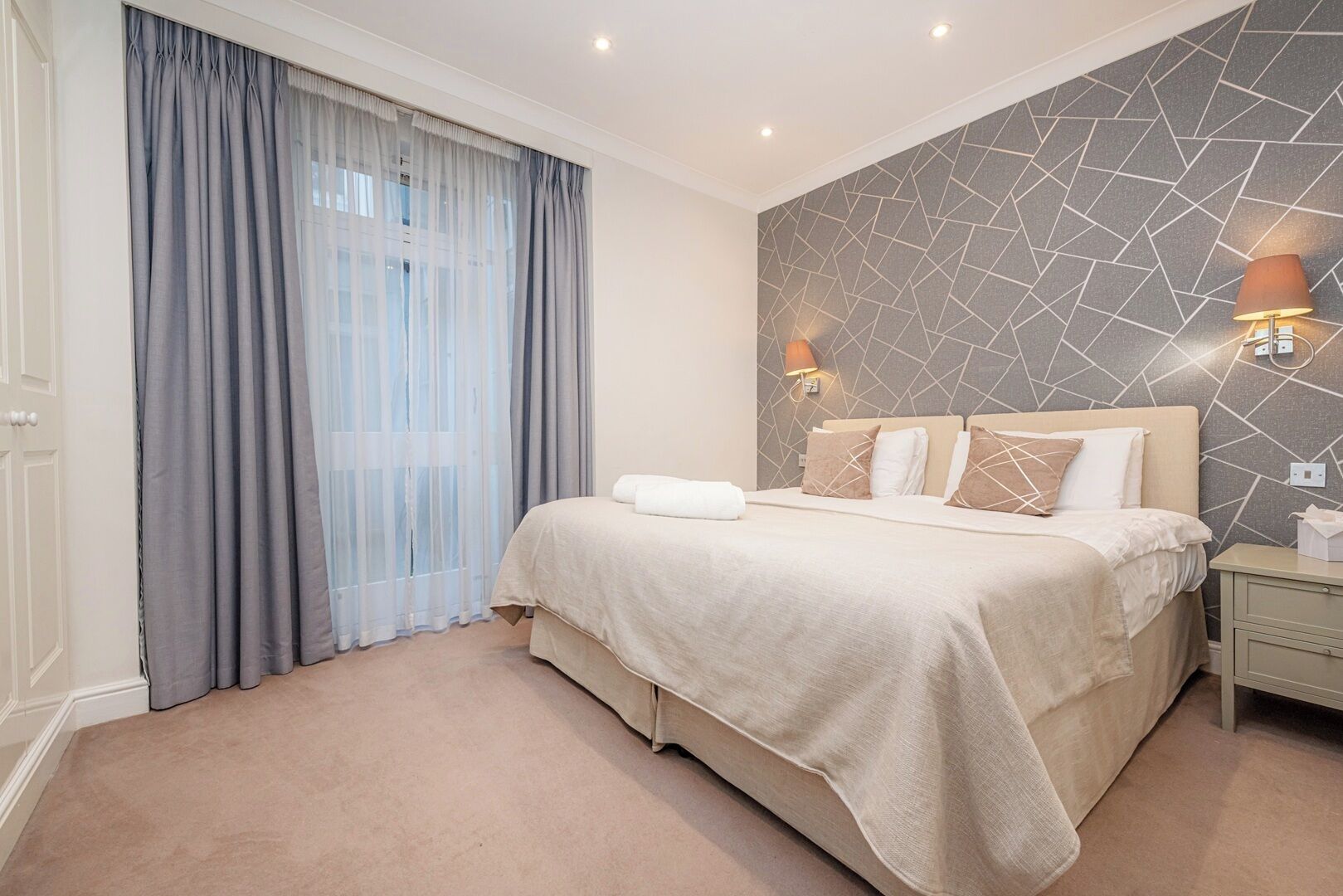 Quarto de hotel com cama, cortinas, uma parede com padrão geométrico e mesa de cabeceira com abajur.