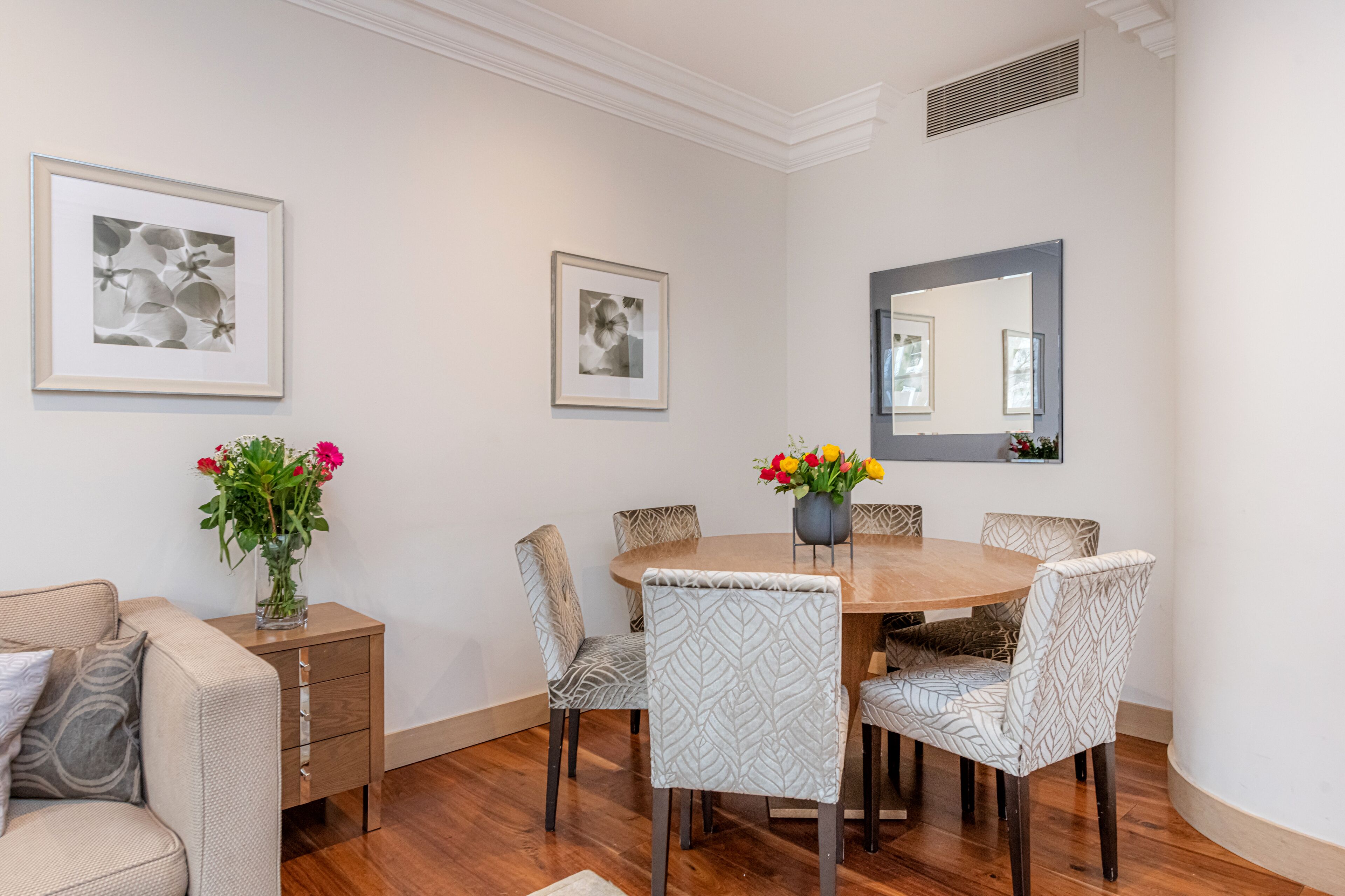 Uma sala de jantar com uma mesa redonda, seis cadeiras estampadas, um vaso com flores e quadros nas paredes.