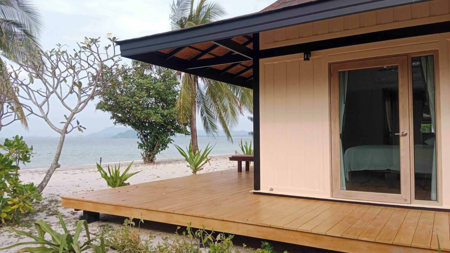 Um deck de madeira com uma porta de vidro que dá para um quarto, com vista para uma praia com palmeiras e o mar.
