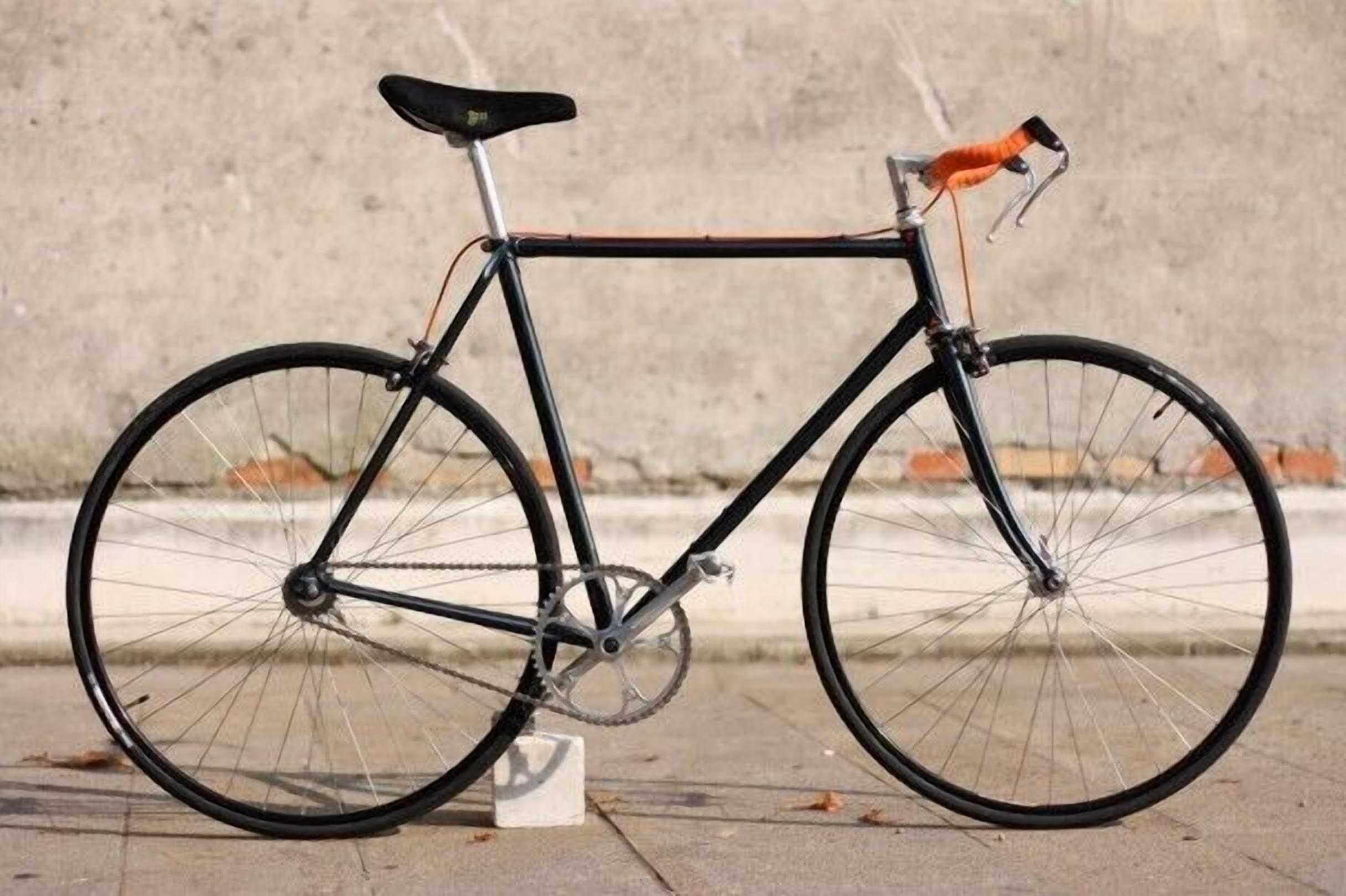 Uma bicicleta ergométrica com estrutura preta e punhos laranja, posicionada sobre uma superfície de concreto diante de uma parede lisa.